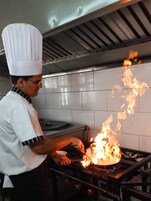 Um chef com uniforme branco e chapéu alto cozinha em um fogão com chamas.