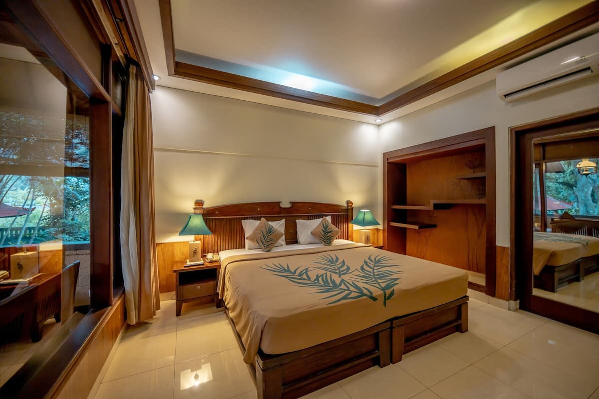 Quarto de hotel com uma cama grande, cabeceira de madeira, luminárias laterais, mesa de cabeceira e vista para o exterior através de uma porta de vidro.