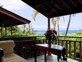 Um deck de madeira com uma mesa e cadeiras, com vista para uma paisagem tropical com palmeiras e o oceano.