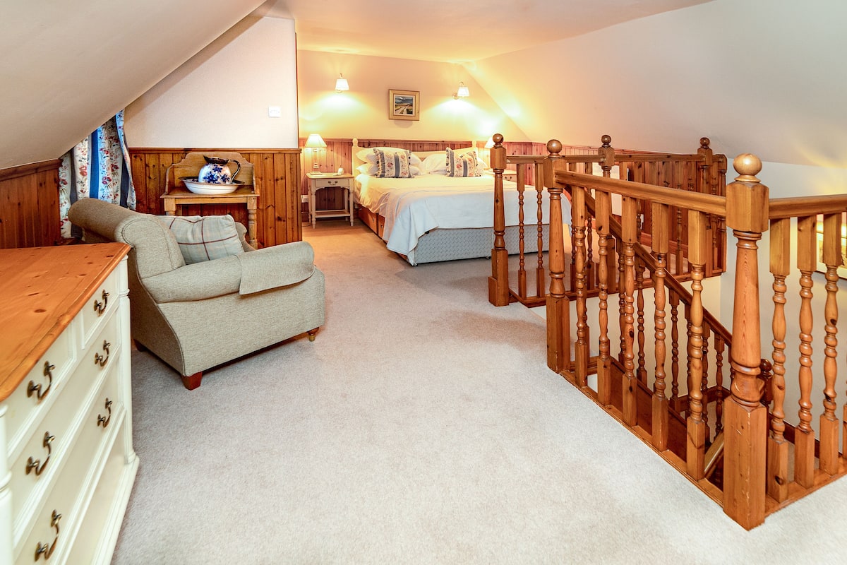 Quarto com escada de madeira, uma cama, mesa de cabeceira, cadeira e cômoda.