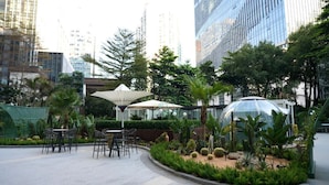 Uma praça urbana ao ar livre com áreas de estar, uma estrutura de estufa transparente e vegetação ao redor.