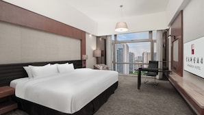 Quarto de hotel com uma cama grande, uma mesa, uma cadeira e vista para a cidade pela janela.