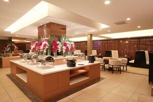 Um lobby de hotel moderno com uma ilha central que inclui um serviço de buffet, área de estar com sofás e cadeiras, e um grande arranjo de flores.