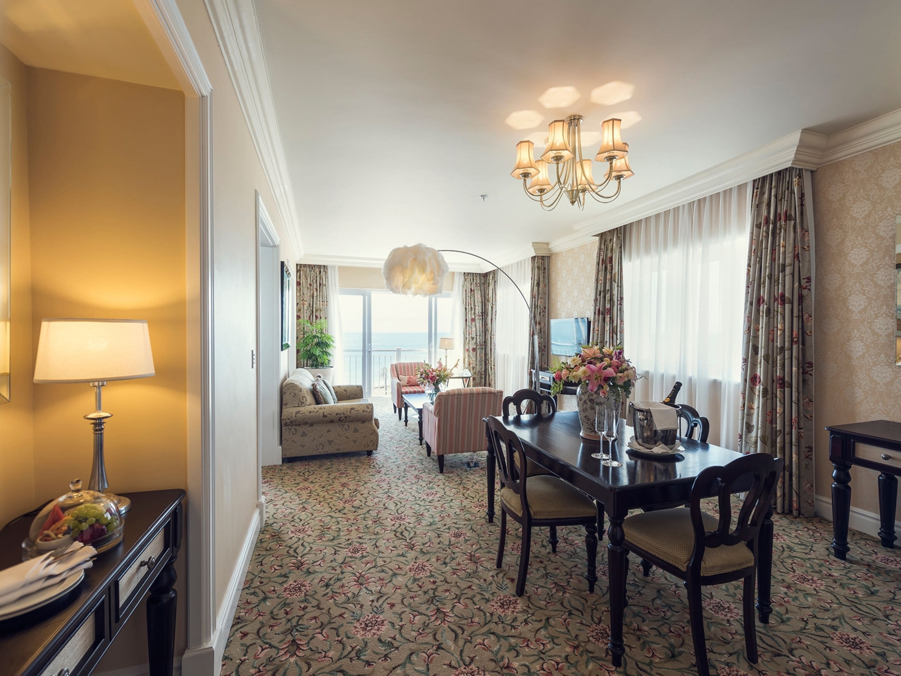 Quarto de hotel com área de refeições, sofá, lustre e vista para o mar.