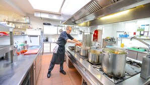 Uma cozinha profissional com eletrodomésticos de aço inoxidável, um chef preparando alimentos e diversos utensílios e ingredientes de cozinha.
