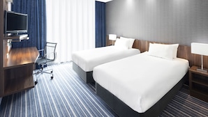 Quarto de hotel com duas camas, uma escrivaninha com televisão, uma cadeira e uma janela grande com cortinas.