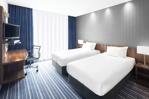 Quarto de hotel com duas camas, uma escrivaninha com televisão, uma cadeira e uma janela grande com cortinas.