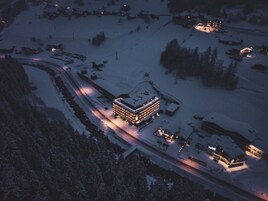 Paisagem coberta de neve com um edifício grande, com várias janelas e telhado plano, rodeado por árvores carregadas de neve e algumas estruturas menores.