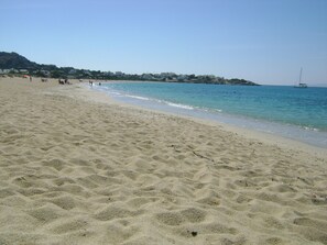 Uma praia arenosa com água azul cristalina, algumas pessoas e edifícios ao fundo.