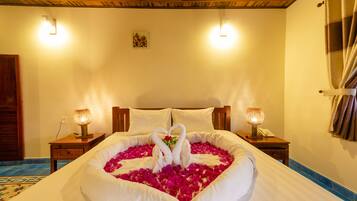 Cama bem arrumada com arranjo de pétalas de rosa em formato de coração e dois cisnes, com duas luminárias laterais e uma mesa de cabeceira com telefone.