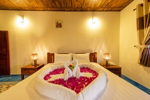 Cama bem arrumada com arranjo de pétalas de rosa em formato de coração e dois cisnes, com duas luminárias laterais e uma mesa de cabeceira com telefone.