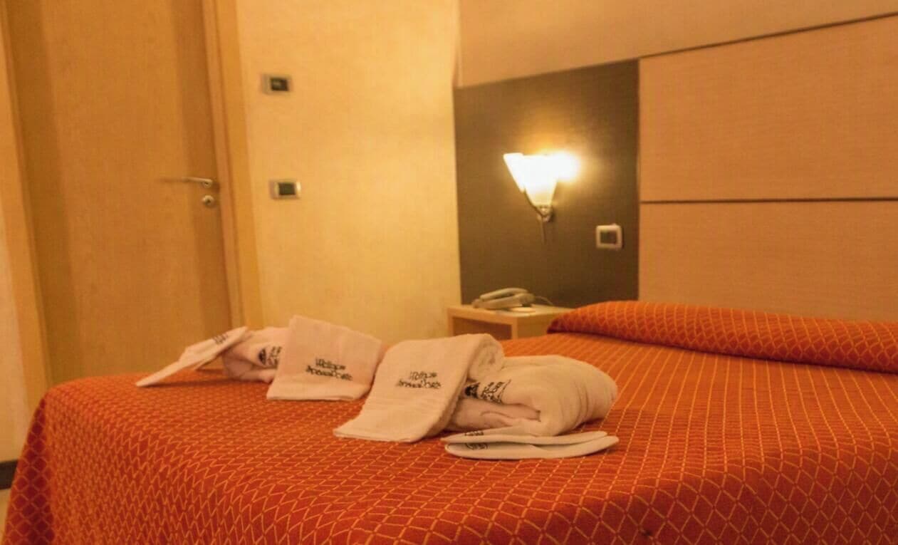 Quarto de hotel com cama, mesa de cabeceira, luminária na parede e porta.