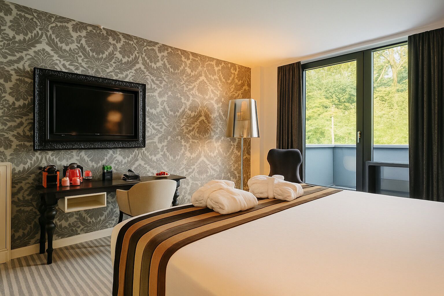 Quarto de hotel com uma cama grande, televisão de tela plana na parede, escrivaninha com abajur e vista para o verde pela janela.