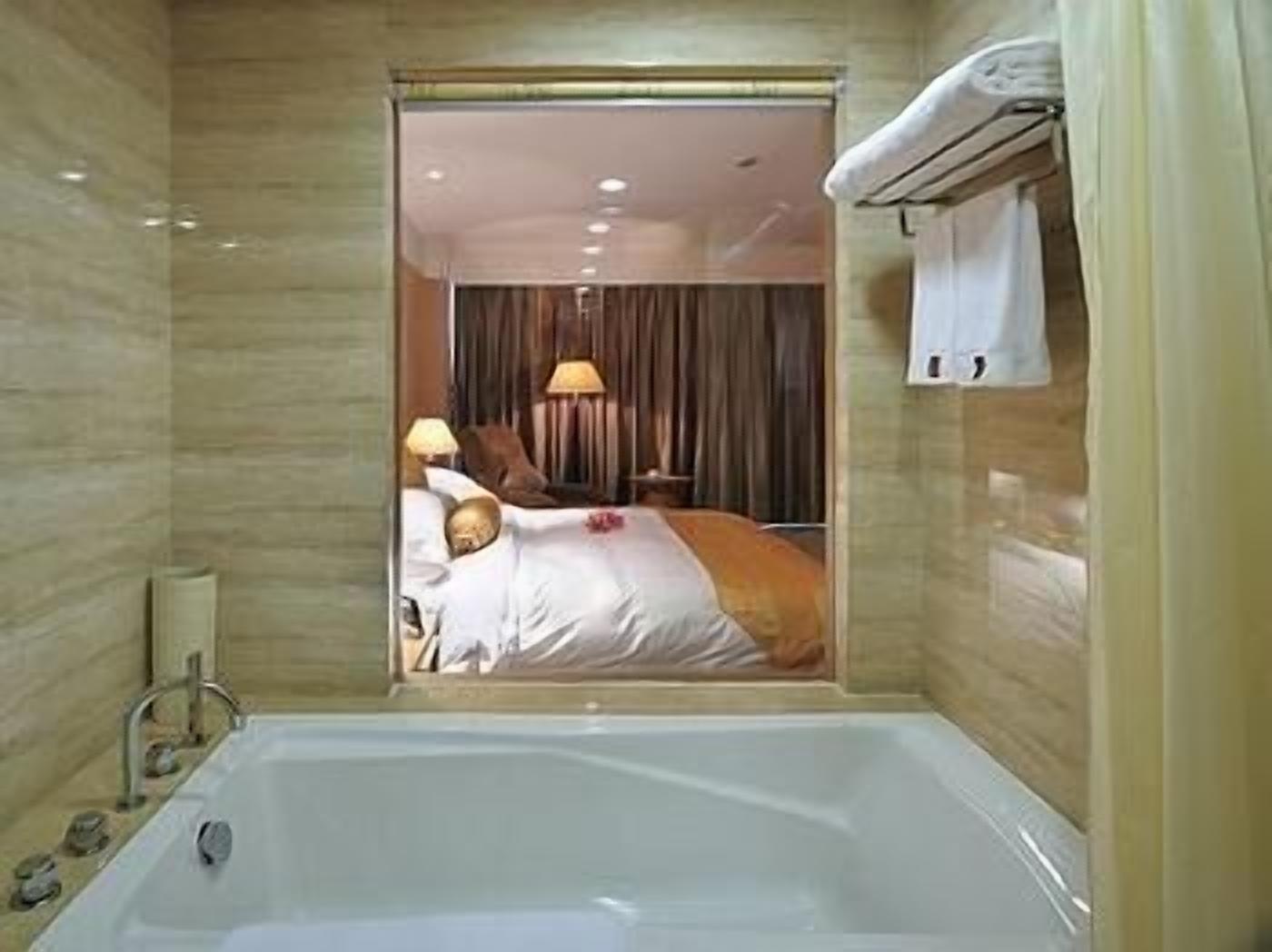 Casa de banho de hotel com uma banheira grande, zona de duche e uma cama visível com cortina.