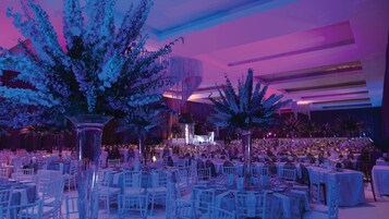 Um espaço para eventos formais, com mesas e cadeiras redondas, decorado com arranjos florais e um palco ao fundo.