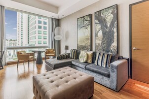 Uma sala de estar moderna com um sofá seccional cinza, uma mesa de centro redonda e um banquinho estofado. Há duas grandes fotos emolduradas na parede, uma porta de madeira e vista de um edifício através de uma grande janela.