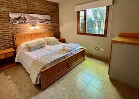 Um quarto com cama de madeira, mesa de cabeceira, janela com vista para árvores e parede de tijolos com um quadro.