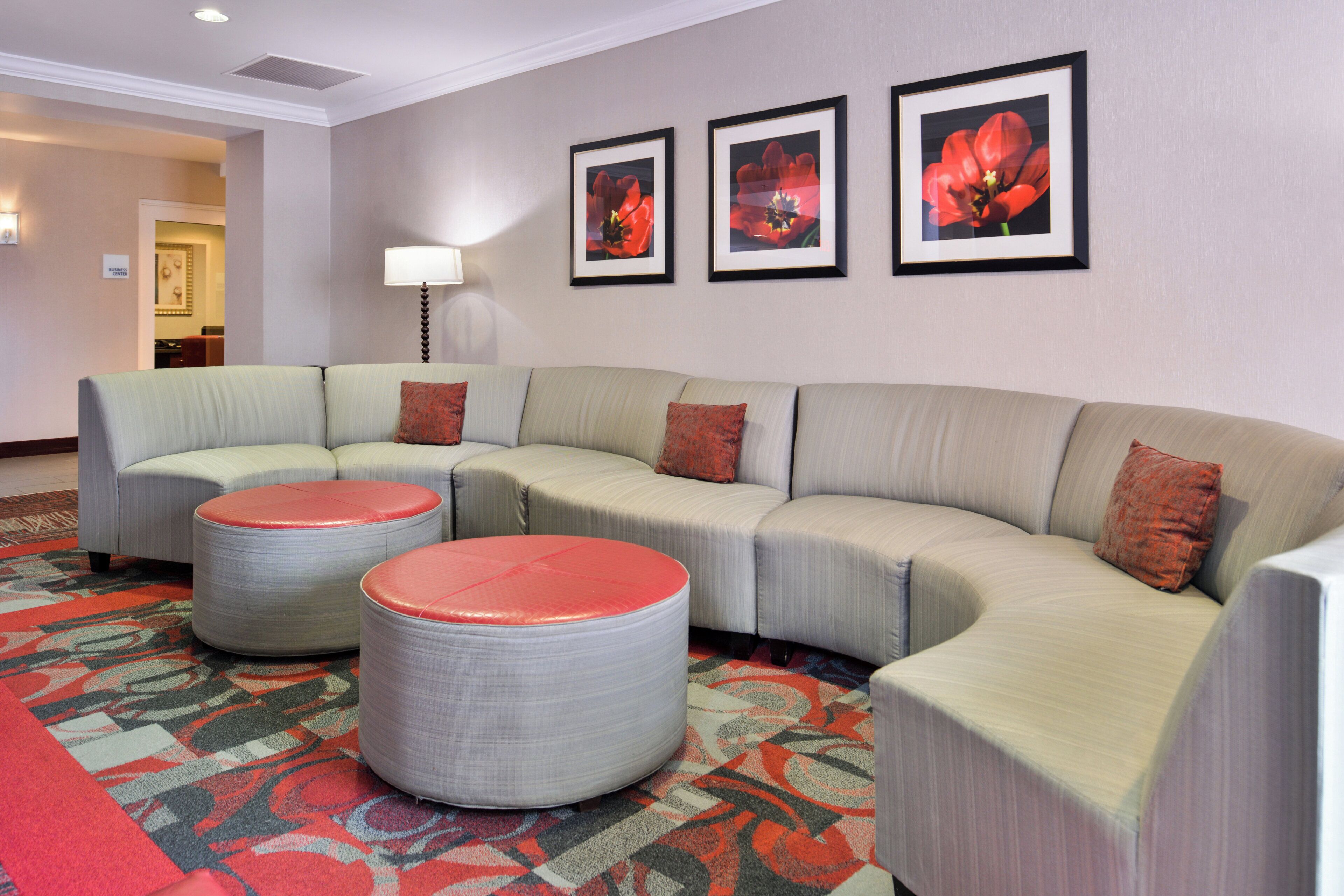 Um hall de hotel com um sofá em forma de L, dois banquinhos, um tapete estampado e três obras de arte florais emolduradas na parede.