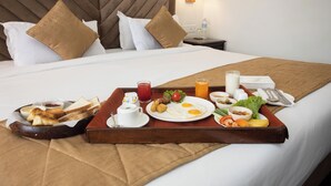 Cama bem arrumada com cabeceira marrom, almofadas brancas e bandeja com itens de café da manhã, incluindo torradas, ovos e suco.