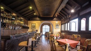 Uma área de bar com banquetas de madeira e um balcão comprido exibindo diversos frascos. Há mesas com cadeiras ao fundo, e uma grande janela em arco é visível.