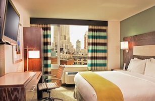 Quarto de hotel com cama, escrivaninha, cadeira e televisão.