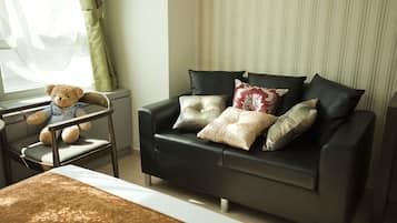 Uma sala de estar moderna com um sofá preto, almofadas decorativas, um urso de pelúcia numa cadeira e uma janela com cortinas leves.