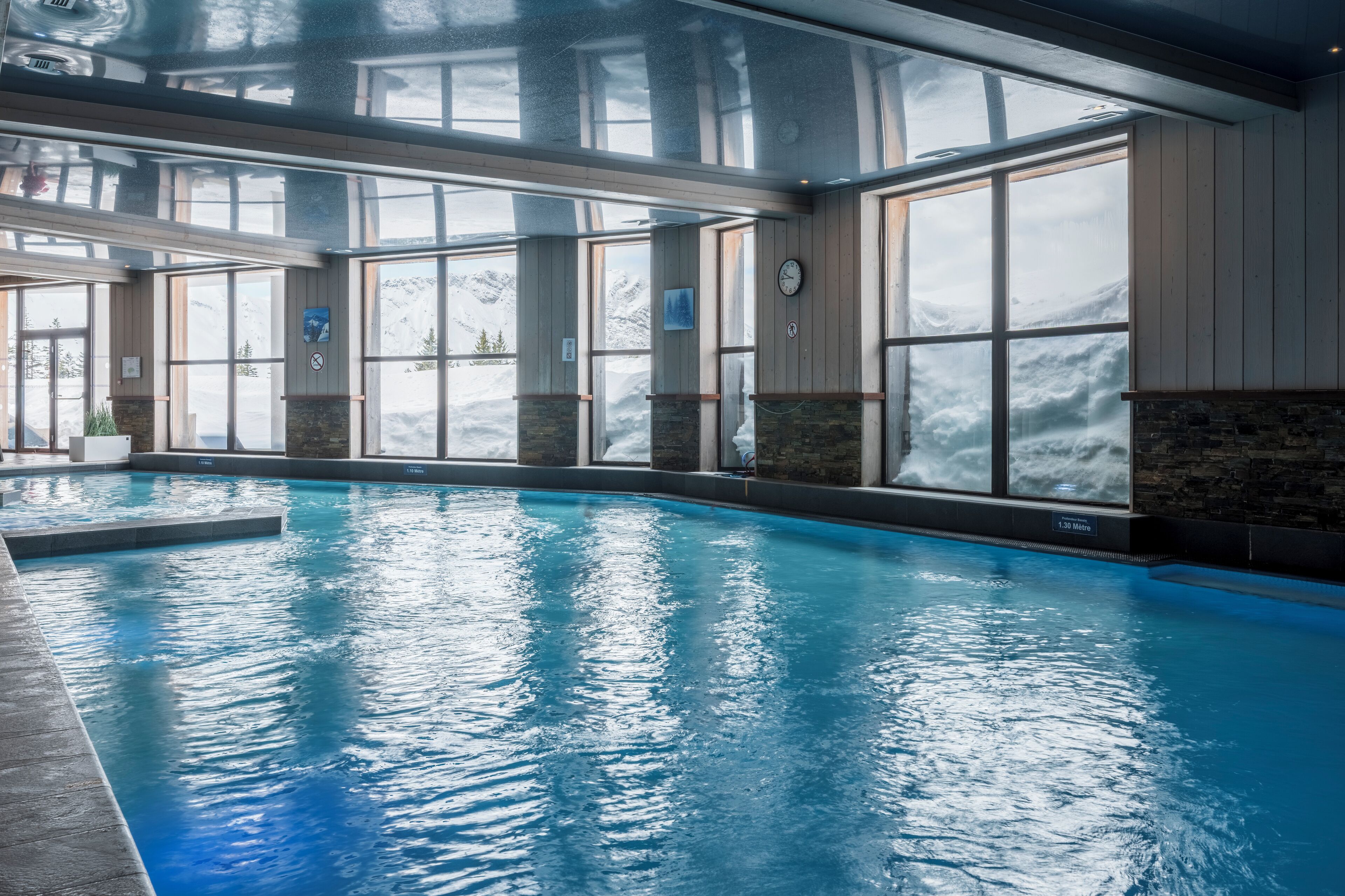 Piscina interior com amplas janelas, paredes de pedra e um relógio na parede.