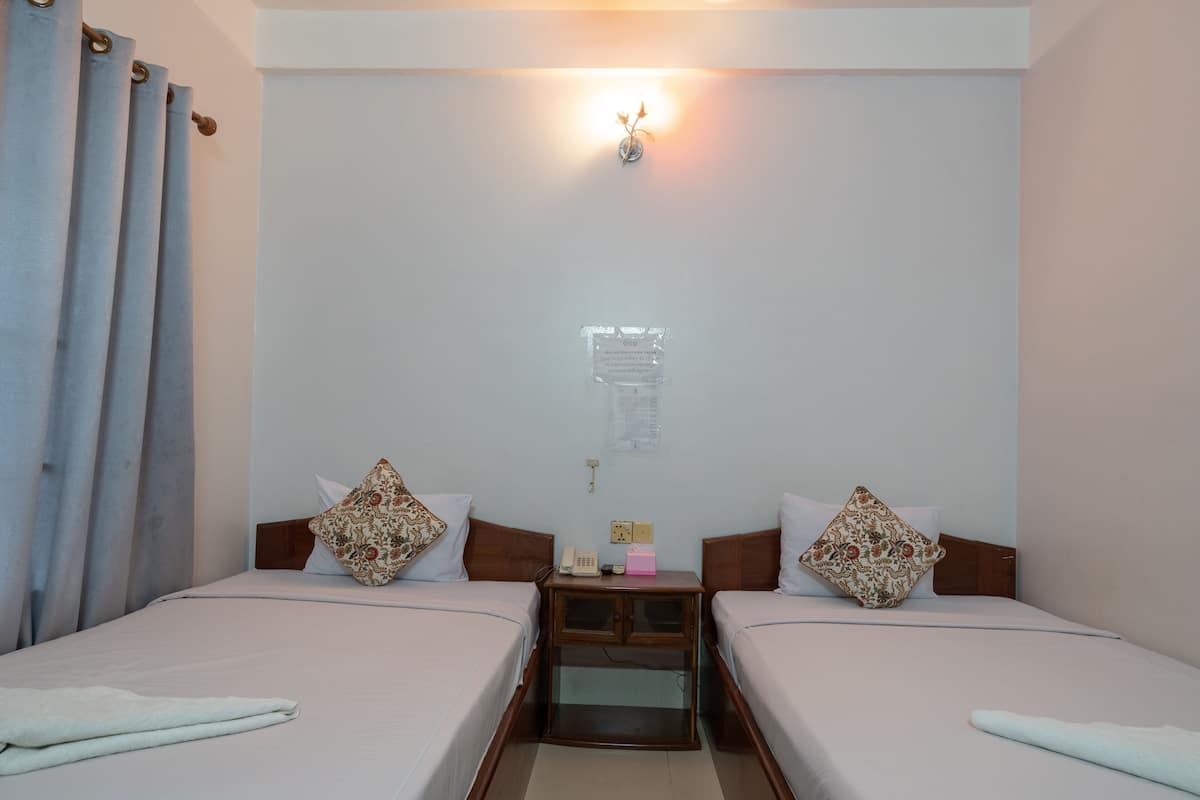 Quarto duplo com duas camas, mesa de cabeceira com luminária, janela com cortinas e luminária na parede.