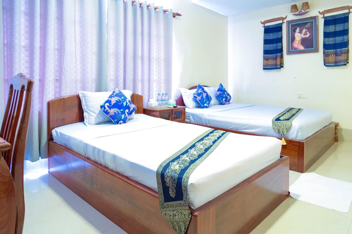 Quarto de hotel com duas camas de madeira, cada uma com colchão branco e almofadas decorativas azuis. Há uma cadeira de madeira e uma mesa pequena com uma luminária.