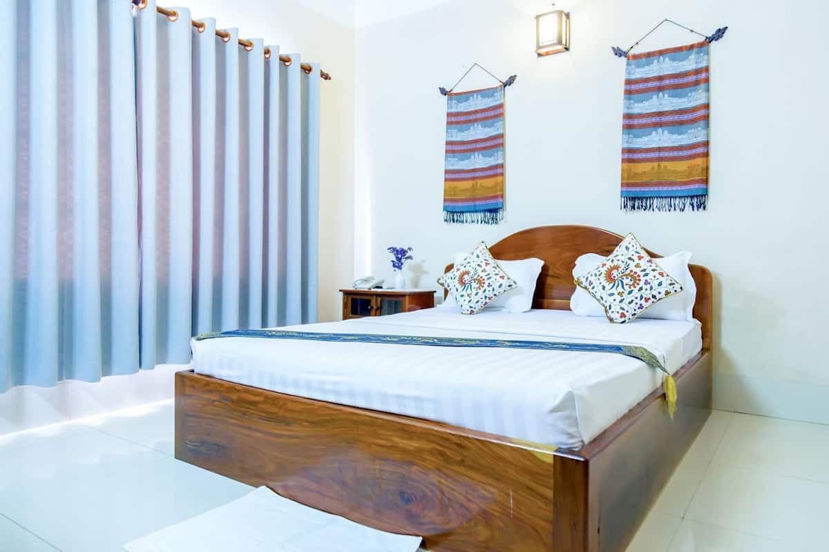 Quarto de hotel com cama de madeira, roupa de cama branca, mesa de cabeceira, luminária e dois objetos decorativos na parede.