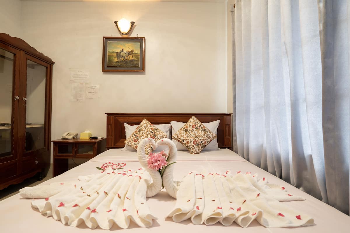 Quarto de hotel com uma cama bem arrumada, um armário de madeira, uma mesinha com telefone e abajur, e um quadro na parede.