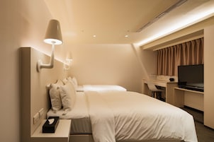 Quarto de hotel com cama, mesa de cabeceira, luminária, escrivaninha, cadeira, televisão e cortinas.