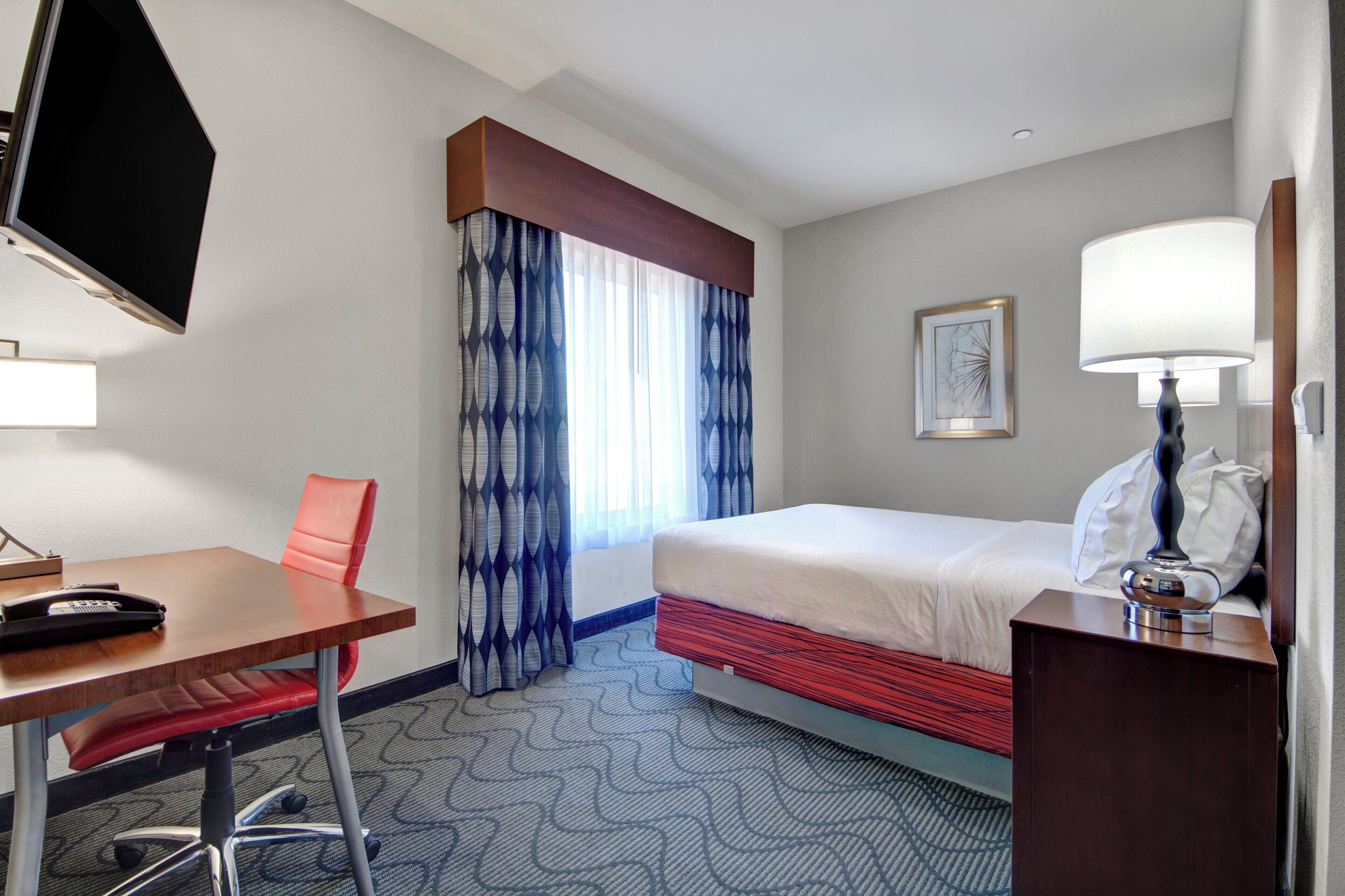 Quarto de hotel com cama, escrivaninha e cadeira, TV fixada na parede, janela com cortinas, luminária de cabeceira e um quadro pendurado na parede.