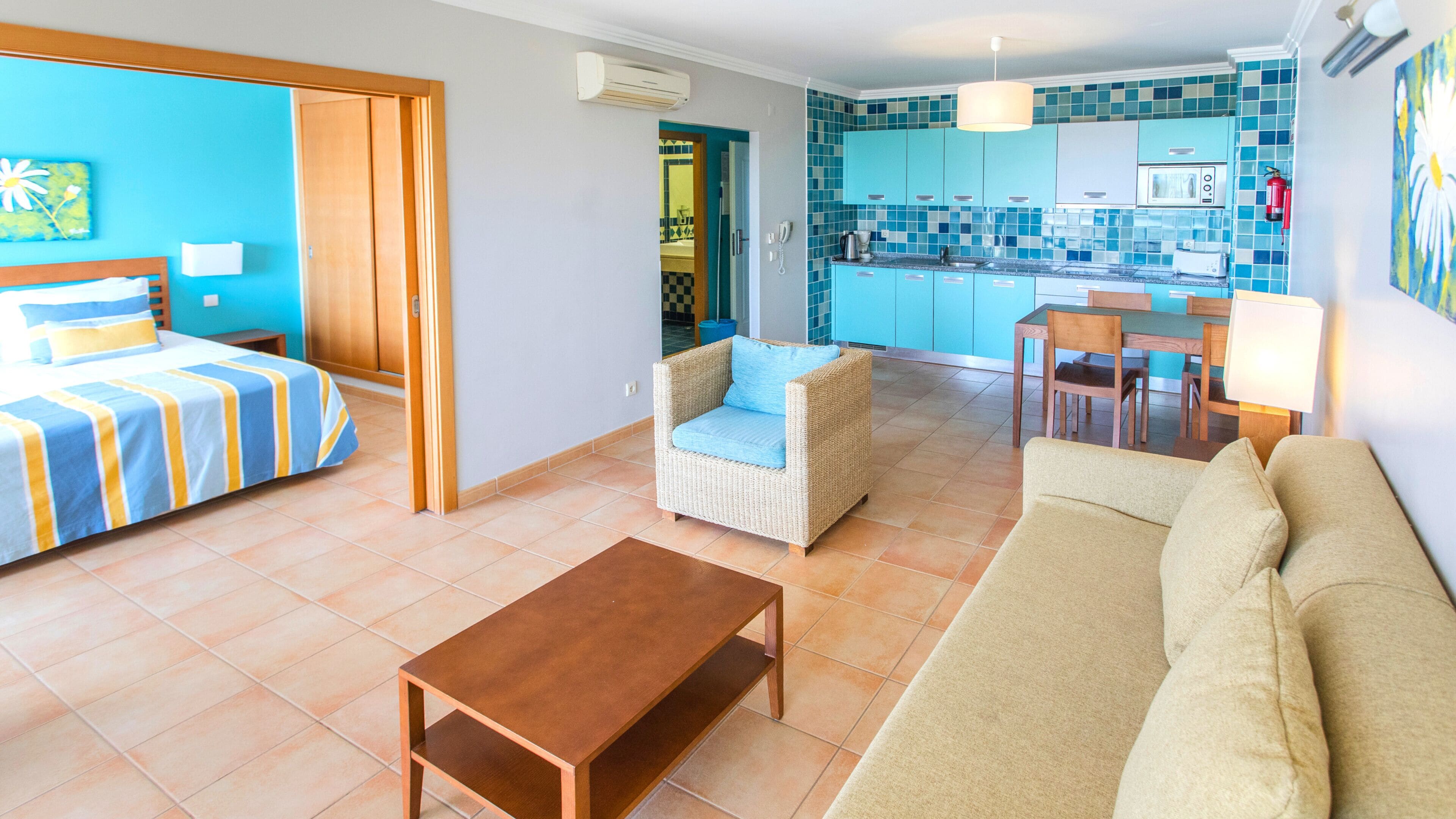 Quarto de hotel moderno com cama, sofá, poltrona, mesa pequena e área de cozinha com mesa de jantar e cadeiras.