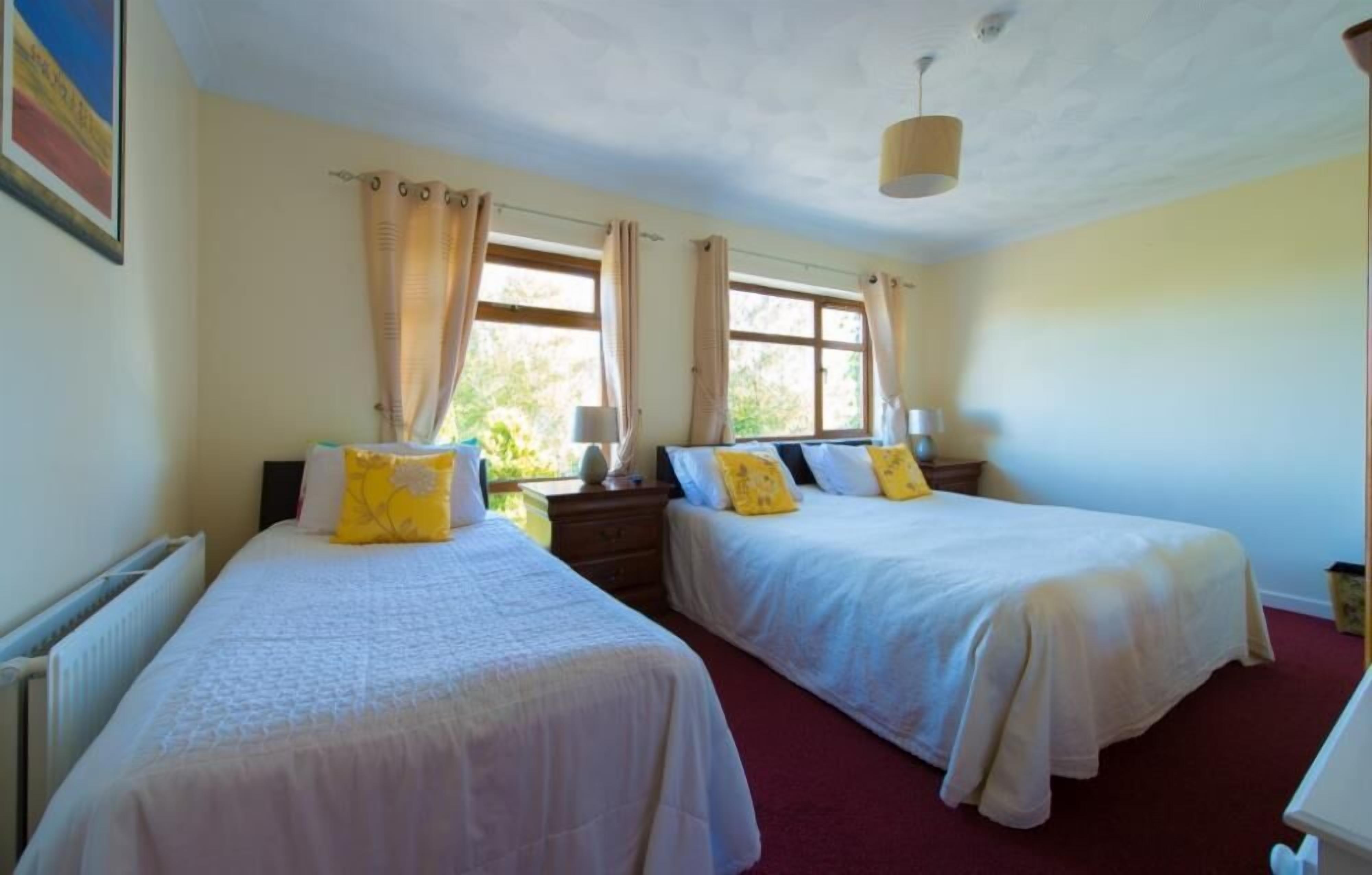 Quarto com duas camas, cortinas bege, janela com vista, radiador e luminária pendente.