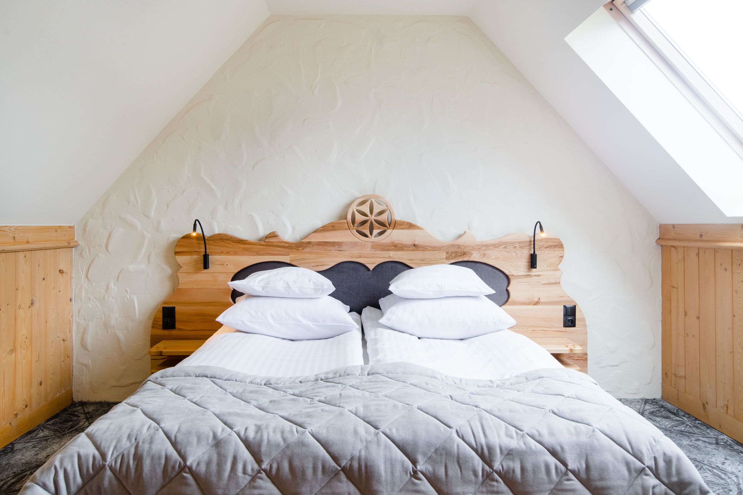 Quarto com cabeceira de madeira, claraboia e cama com colcha cinza.