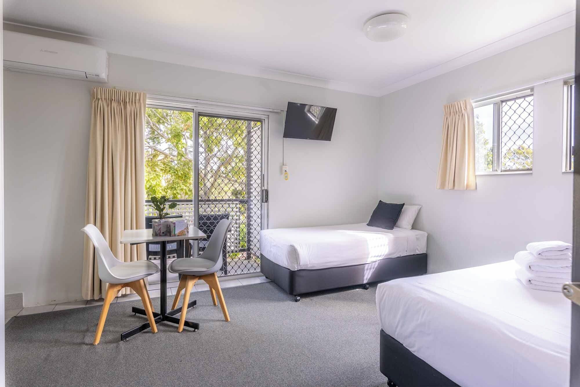 Quarto de hotel com cama, escrivaninha com cadeiras, televisão e janela com cortinas.