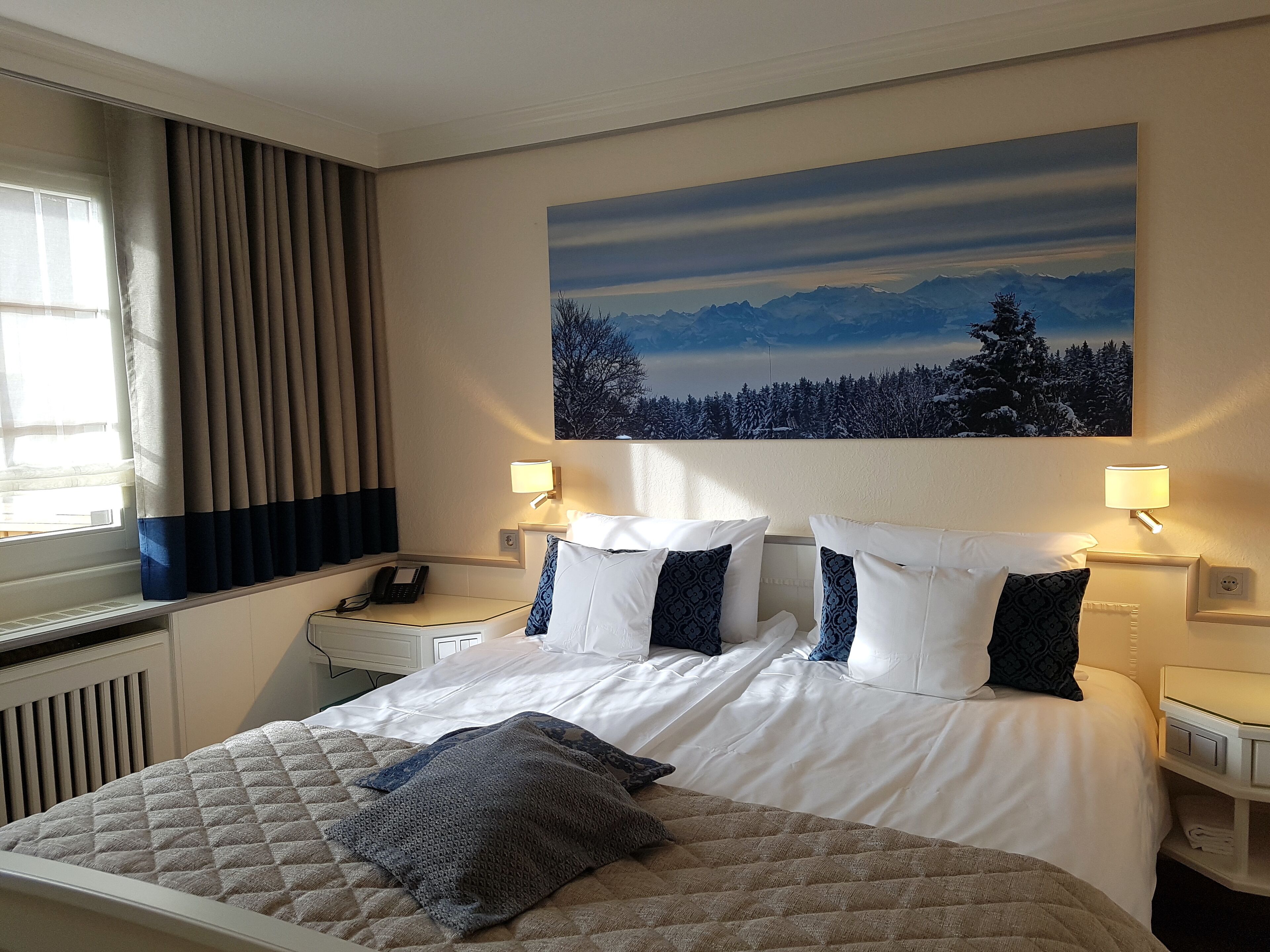 Quarto de hotel com uma cama grande, duas mesas de cabeceira com luminárias, um telefone e um quadro com paisagem de montanha.