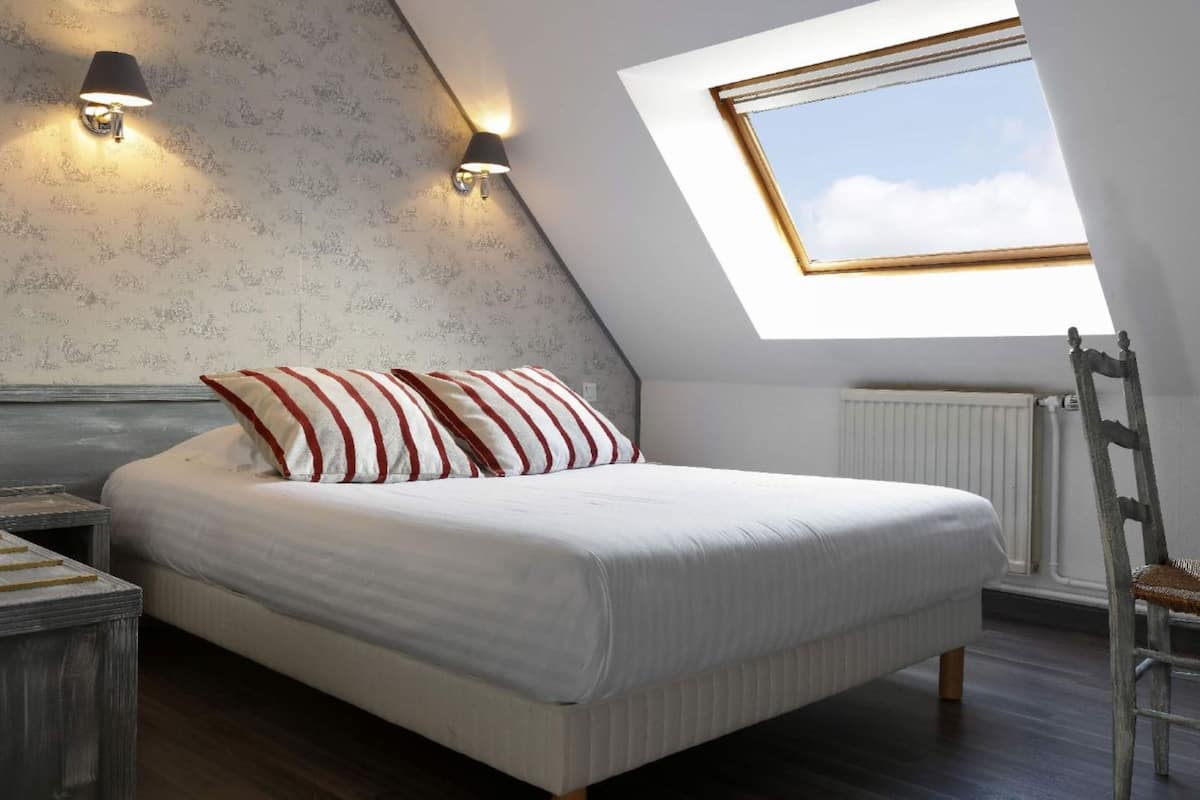 Quarto com teto inclinado, um claraboia, cama com almofadas listradas, duas luminárias fixadas na parede e um radiador.