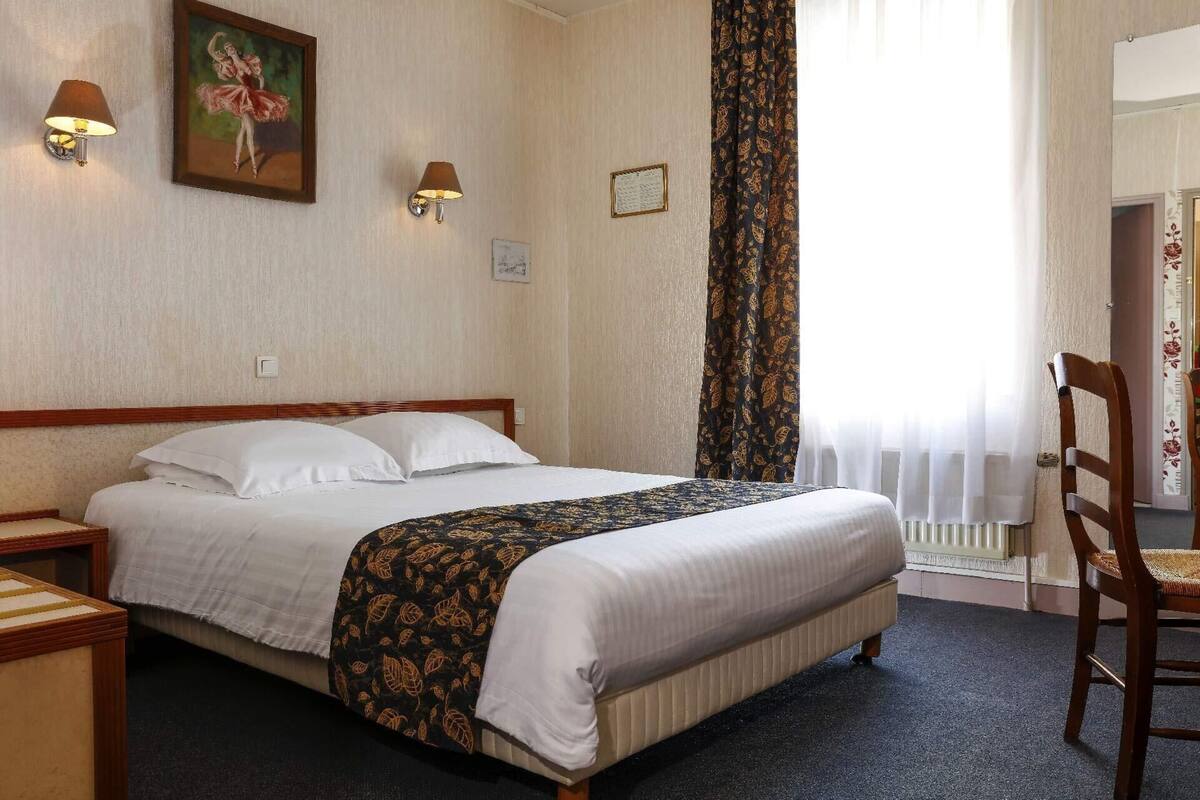 Quarto de hotel com cama, duas luminárias fixadas na parede, um quadro, um certificado emoldurado e uma janela com cortinas.
