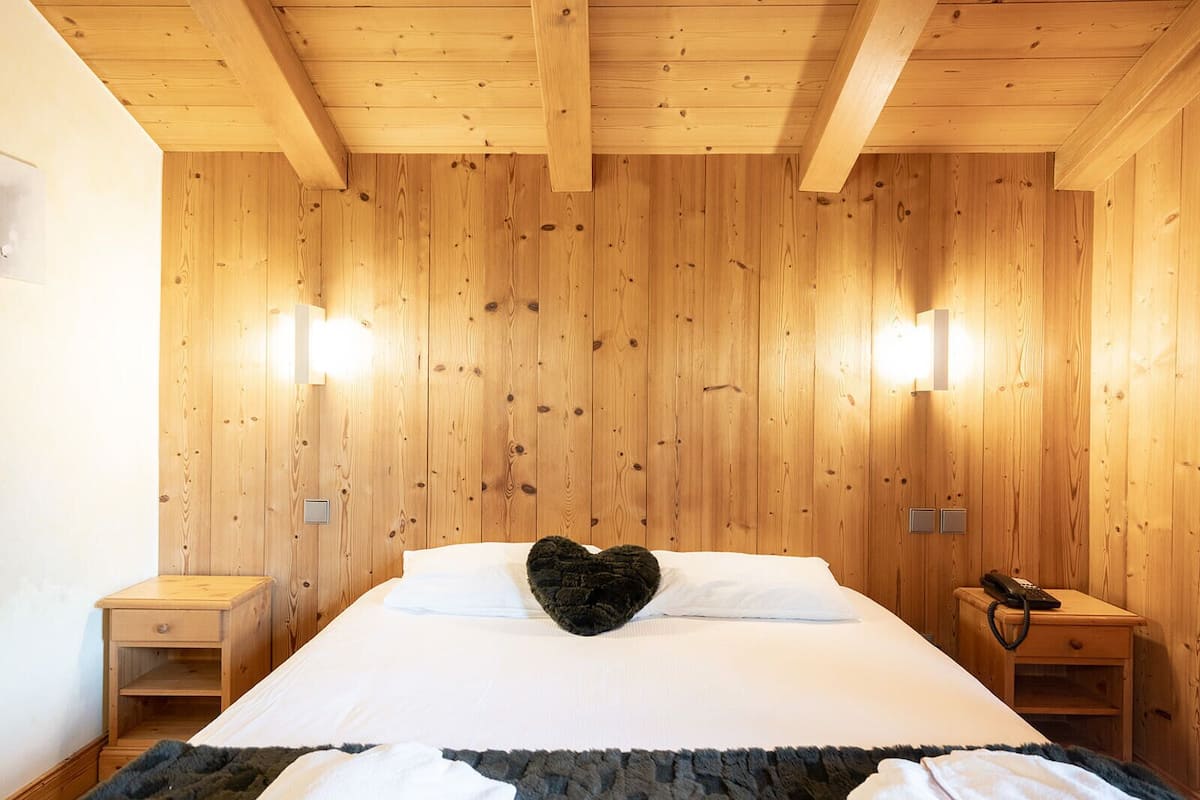 Quarto com painéis de madeira, uma cama, mesas de cabeceira e luminárias fixadas na parede.