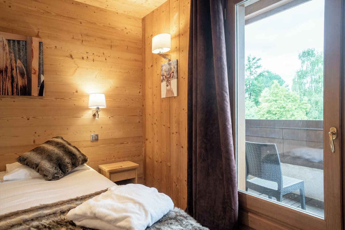 Quarto com painéis de madeira, uma cama, uma mesa de cabeceira, uma luminária e janela com vista para vegetação.