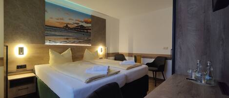 Quarto de hotel moderno com uma cama grande, duas mesas de cabeceira, uma escrivaninha e uma cadeira. Há uma pintura de uma praia na parede.