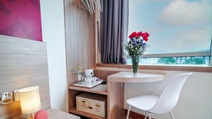 Quarto pequeno e moderno, com uma mesa redonda, uma cadeira branca, um abajur, uma prateleira com artigos de higiene, um vaso com flores e uma janela com vista.