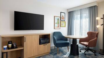 Quarto de hotel com televisão de tela plana, armário de madeira com micro-ondas, duas cadeiras, uma mesa pequena e uma janela grande com cortinas.