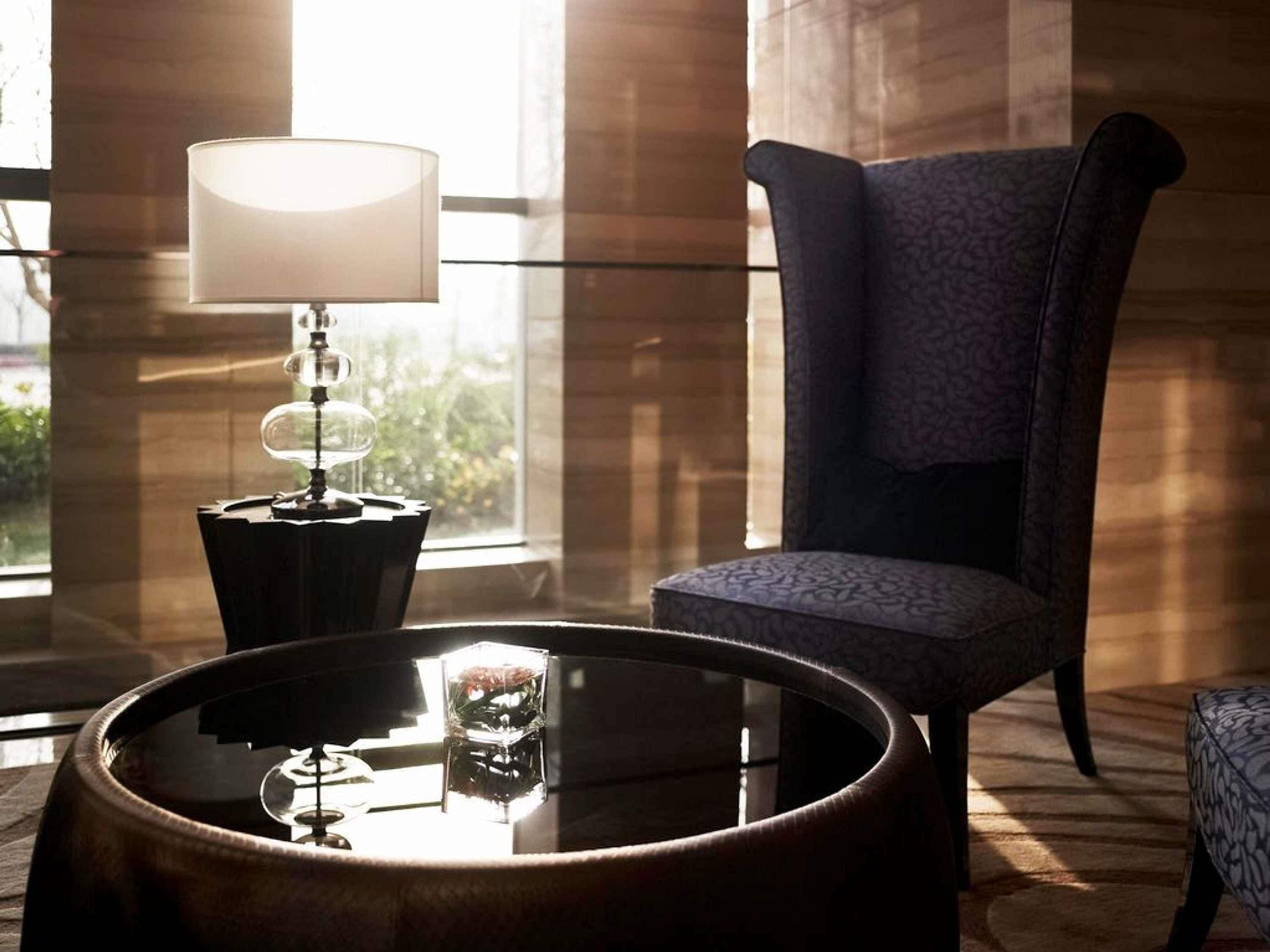 Quarto moderno com uma mesa de centro de vidro redonda, uma poltrona estampada e um abajur sobre uma mesa lateral.