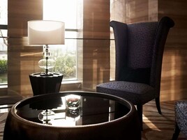 Quarto moderno com uma mesa de centro de vidro redonda, uma poltrona estampada e um abajur sobre uma mesa lateral.