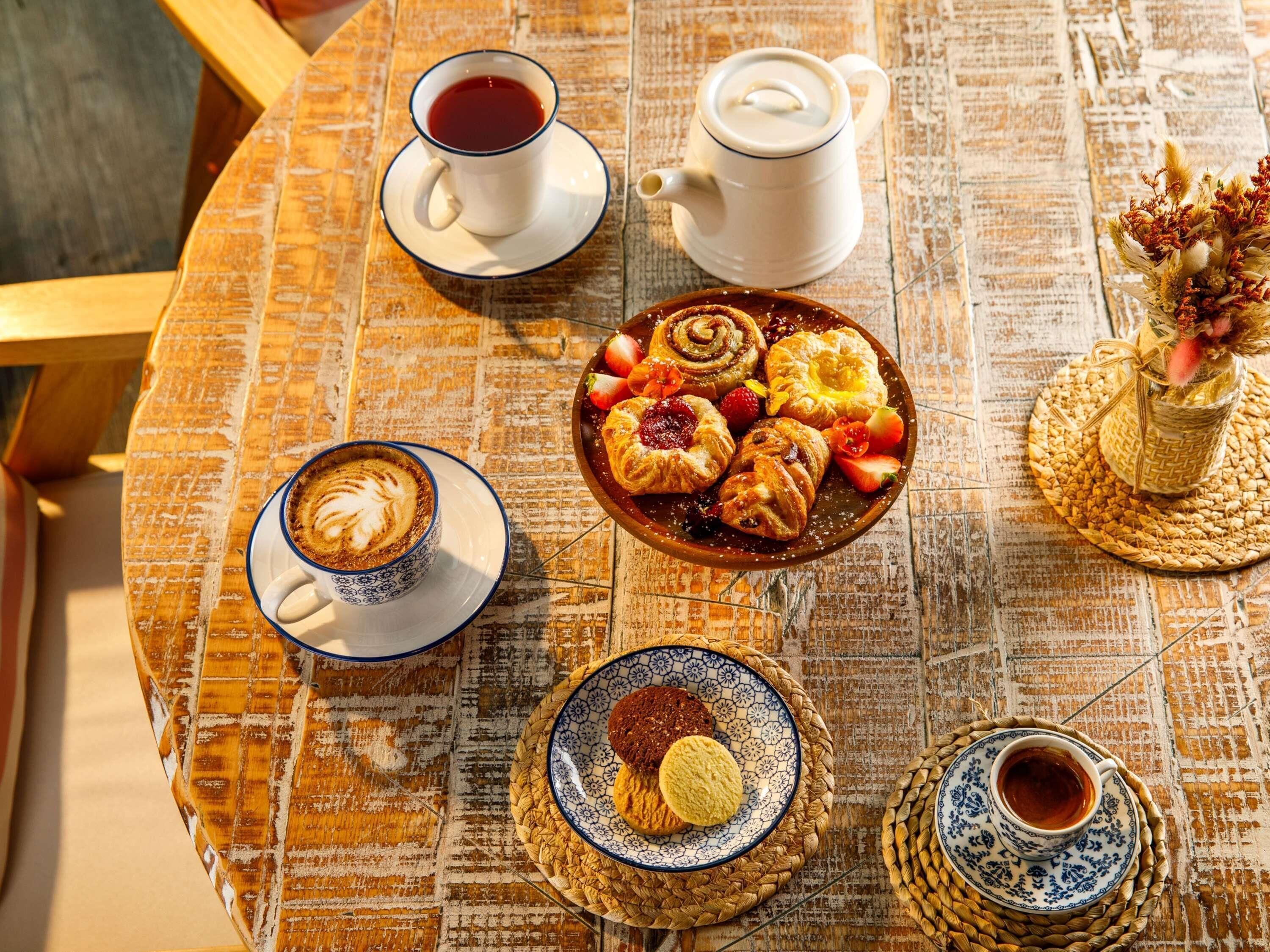 Uma mesa redonda com toalha de madeira rústica, uma chávena de café, um bule, pastelaria e um pequeno vaso com flores secas.