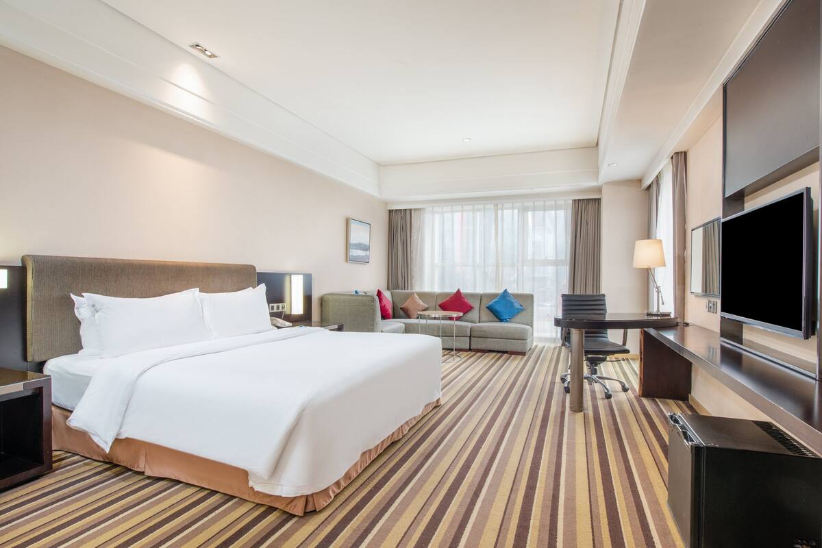 Quarto de hotel com uma cama grande, sofá, escrivaninha, televisão e janela com cortinas.