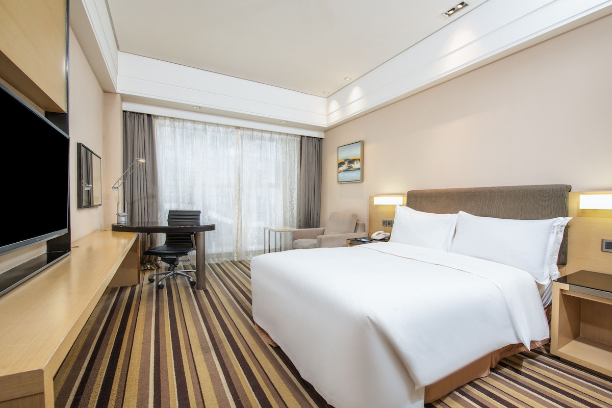 Quarto de hotel com uma cama grande, uma escrivaninha com cadeira, televisão e um quadro na parede.